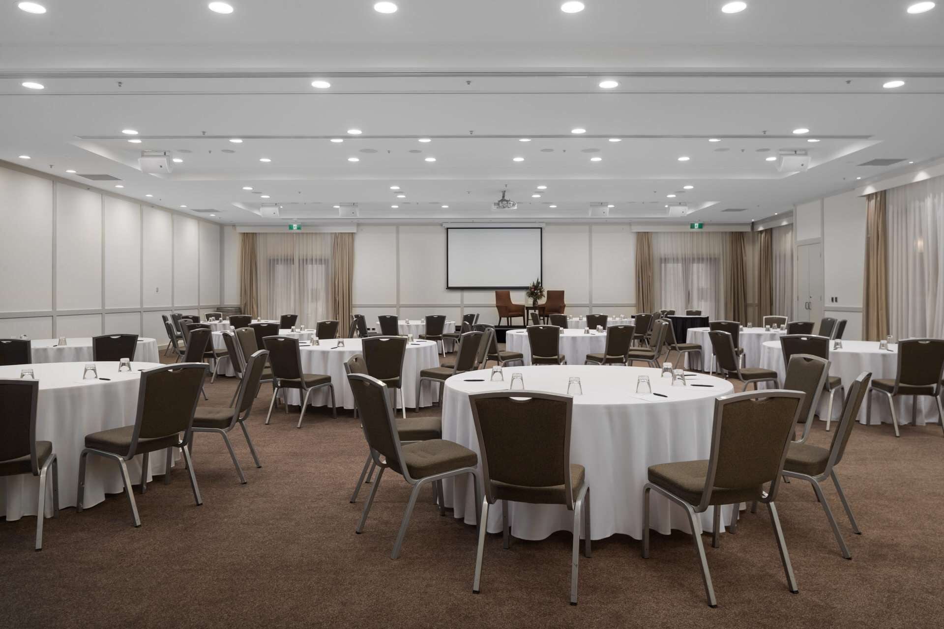 Sala de conferências com mesas redondas, cadeiras, tela de projetor e quadro branco.