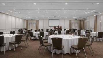 Sala de conferências com mesas redondas, cadeiras, tela de projetor e quadro branco.