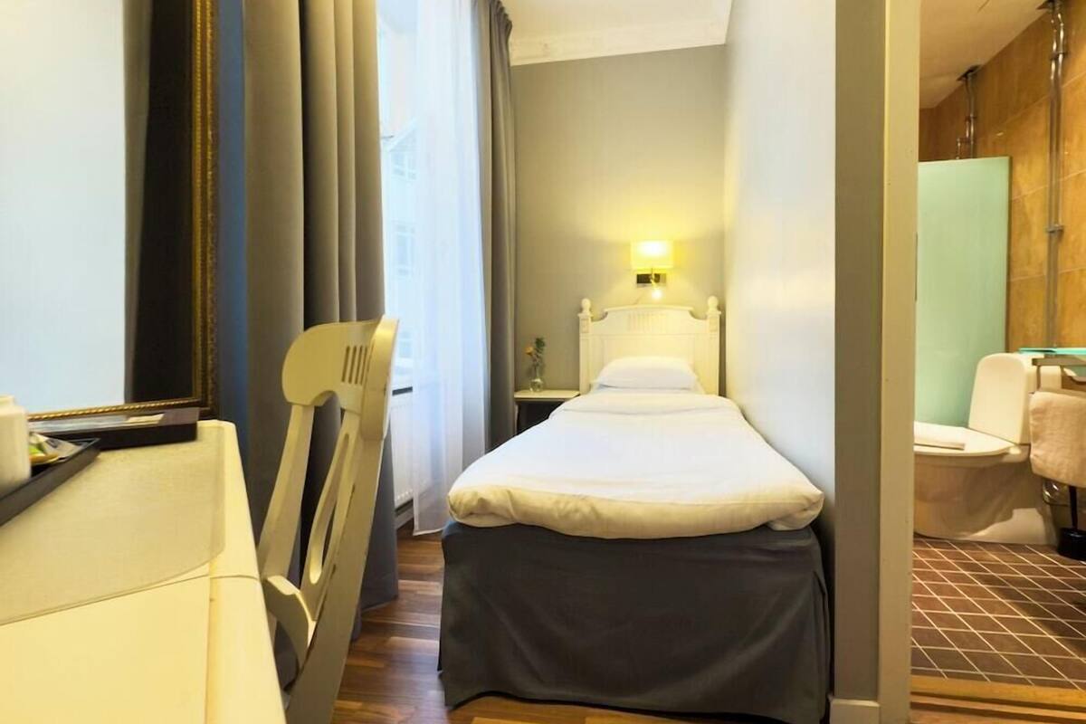 Quarto de hotel com uma cama, uma cadeira, uma mesinha de cabeceira e um banheiro visível através de uma porta aberta.