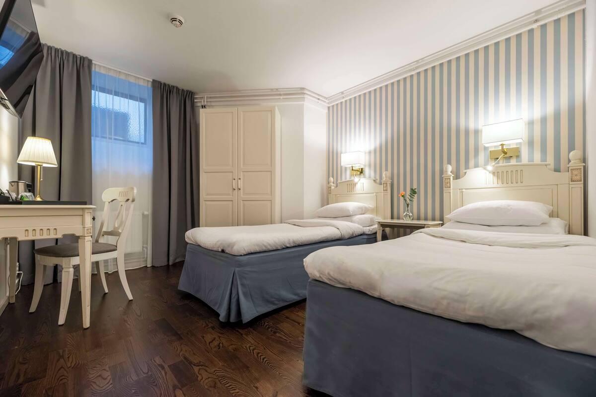 Quarto de hotel com duas camas, uma escrivaninha, uma cadeira e uma janela grande com cortinas.