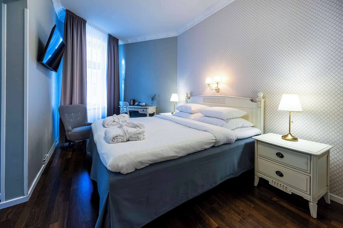 Quarto de hotel com cama, abajures, televisão na parede, escrivaninha com cadeira e janela com cortinas.