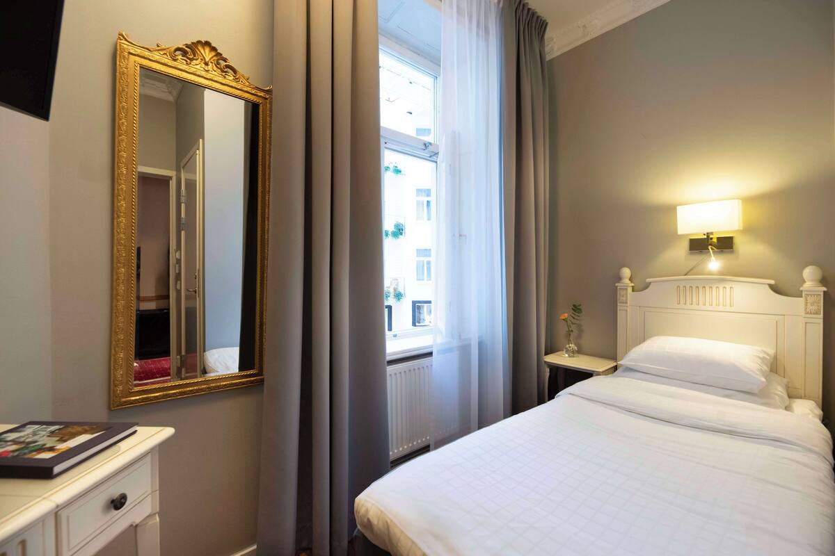 Quarto de hotel com cama, mesa de cabeceira, luminária, espelho e janela com cortinas.