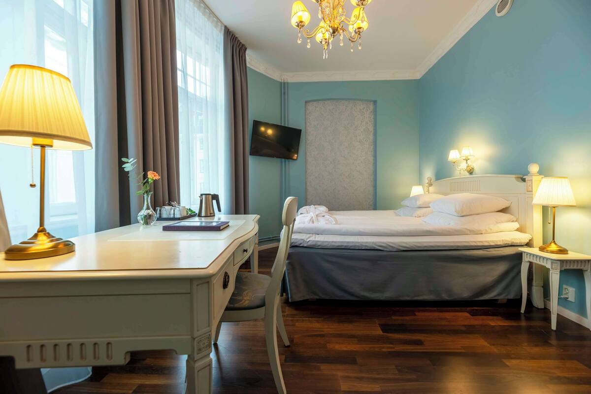 Quarto de hotel com cama, escrivaninha, cadeira, mesas de cabeceira, luminárias, televisão e janela com cortinas.