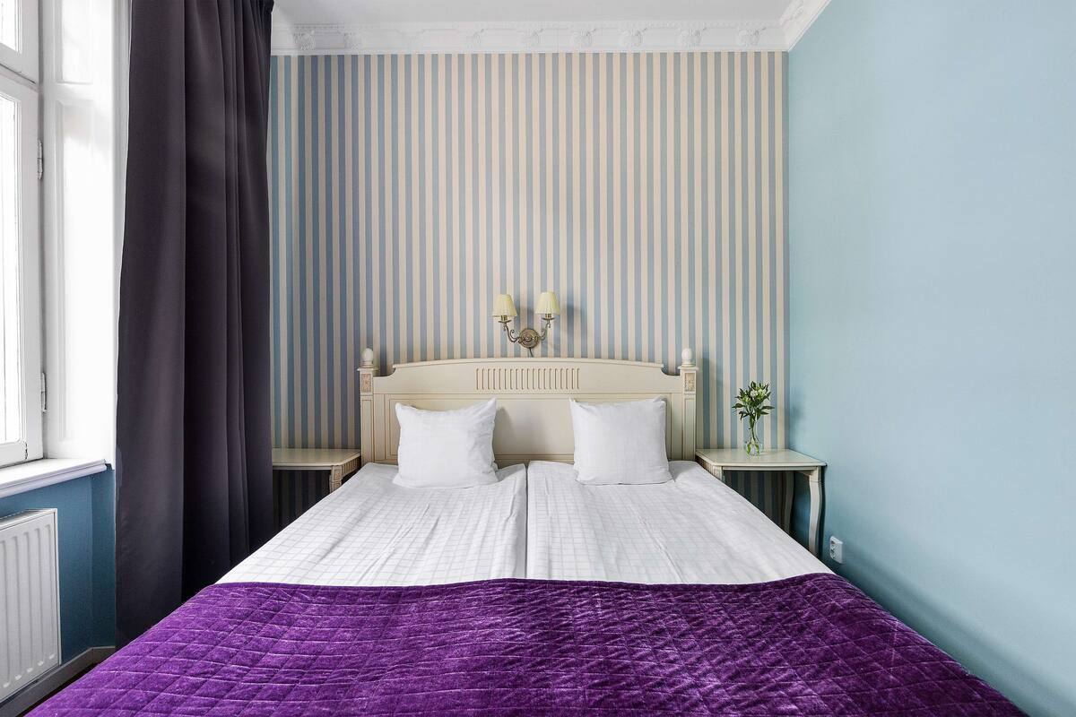 Cama bem arrumada com um edredom roxo, cabeceira branca e parede listrada ao fundo.