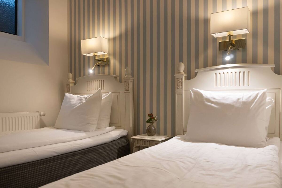 Quarto de hotel com duas camas, papel de parede listrado, luminárias fixadas na parede, radiador e um vaso com flores.