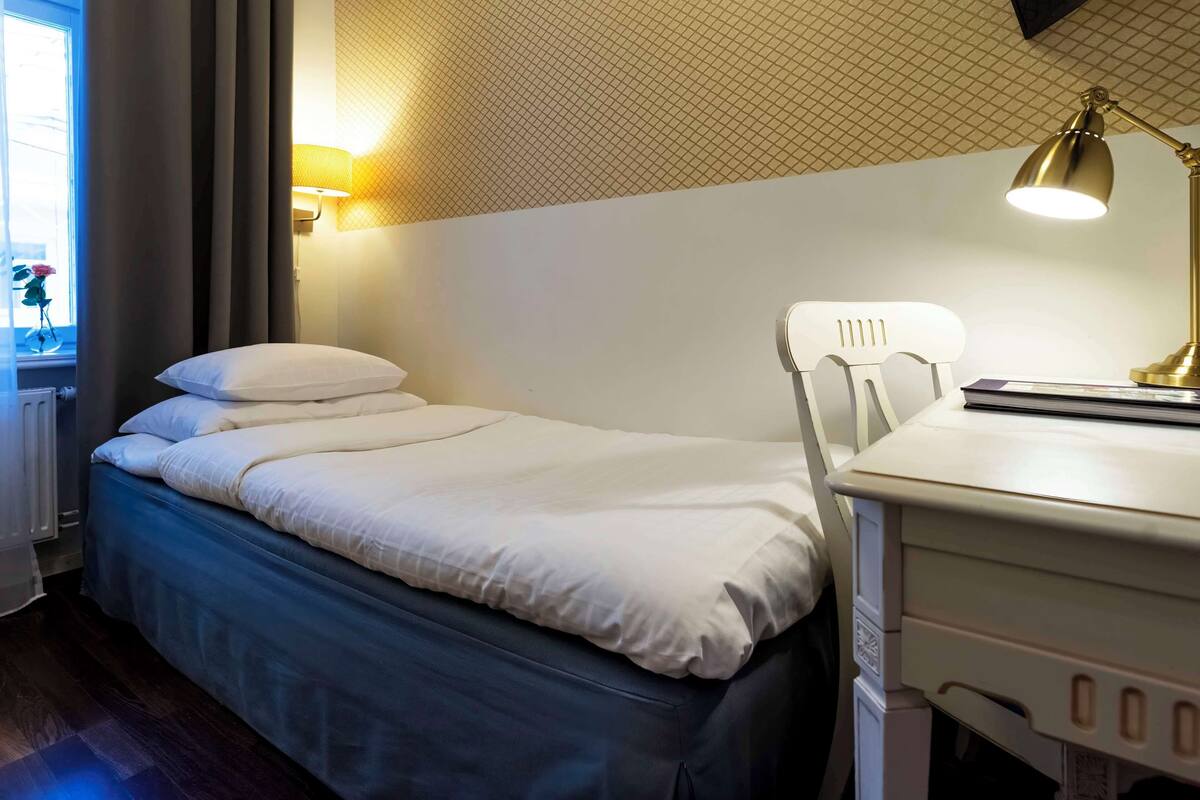 Uma cama de solteiro com roupa de cama branca, uma escrivaninha com candeeiro e uma cadeira num quarto com parede estampada e janela com cortinas.