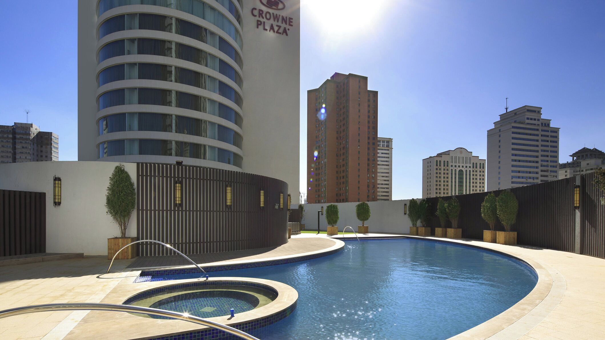 Área da piscina com hidromassagem, cercada por um edifício moderno com a placa 'CROWNE PLAZA'.