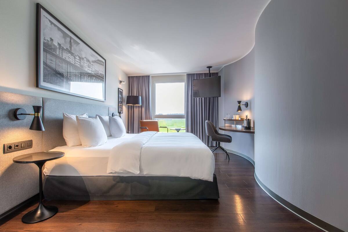 Quarto de hotel moderno com uma cama grande, uma mesinha, televisão e vista para o exterior.