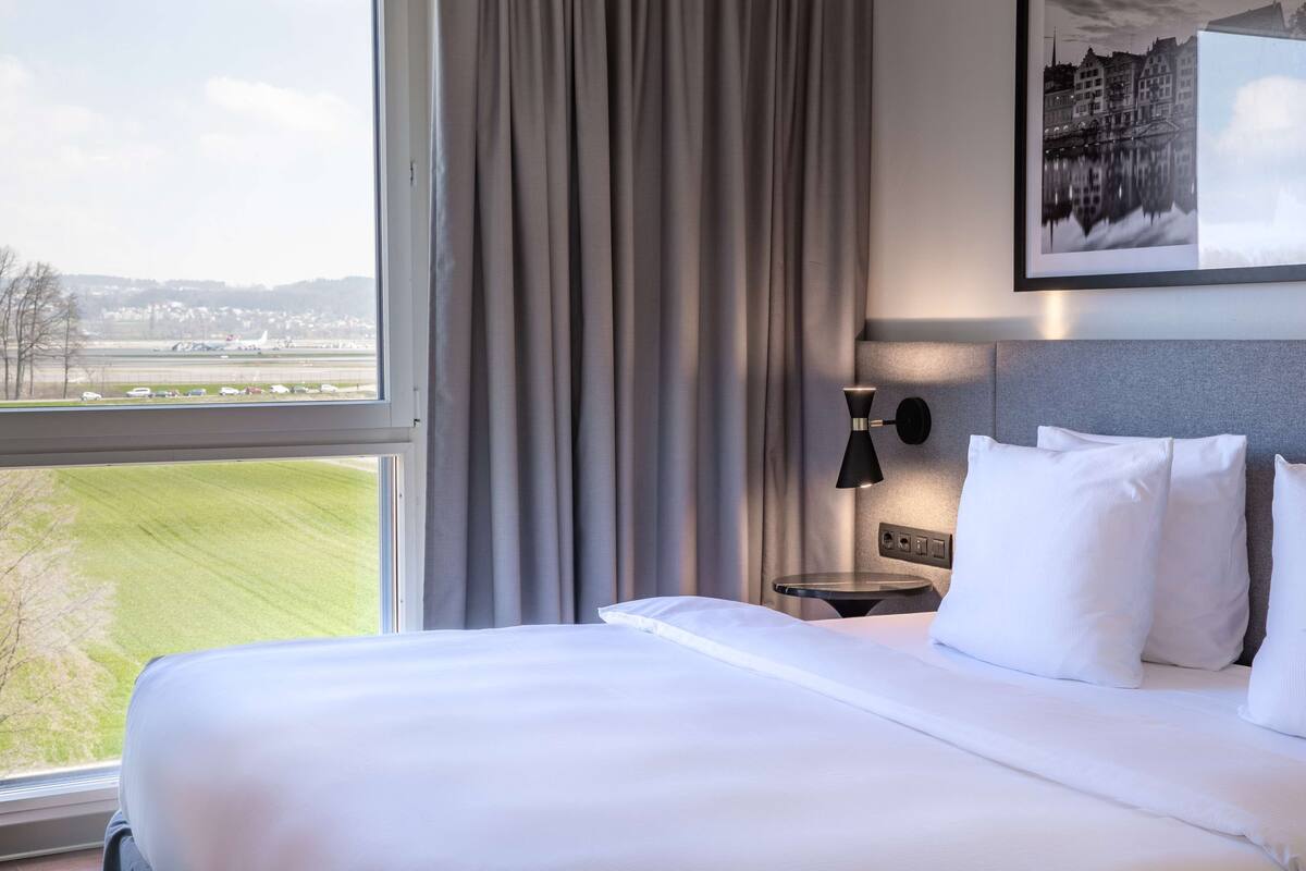 Quarto de hotel com uma cama grande, vista para um campo verde e aeroporto, e uma moldura com uma fotografia na parede.