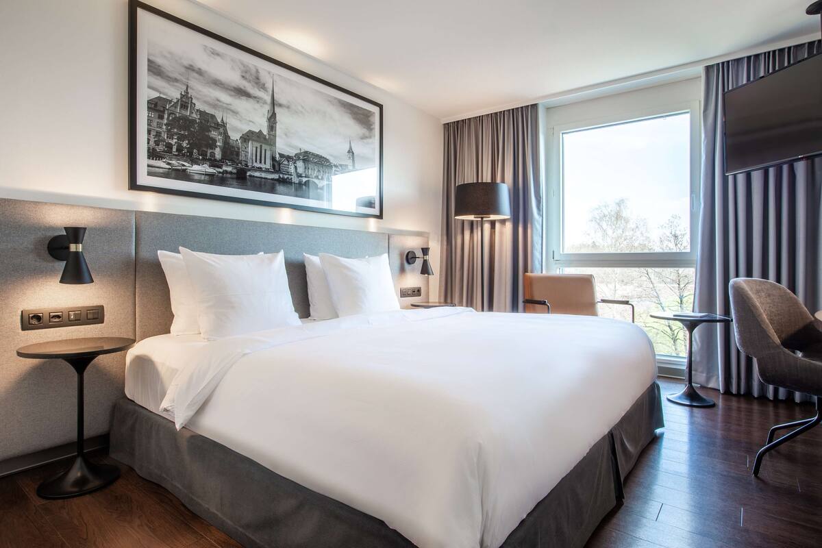 Quarto de hotel com uma cama grande, uma escrivaninha, uma cadeira, televisão e janela com cortinas.