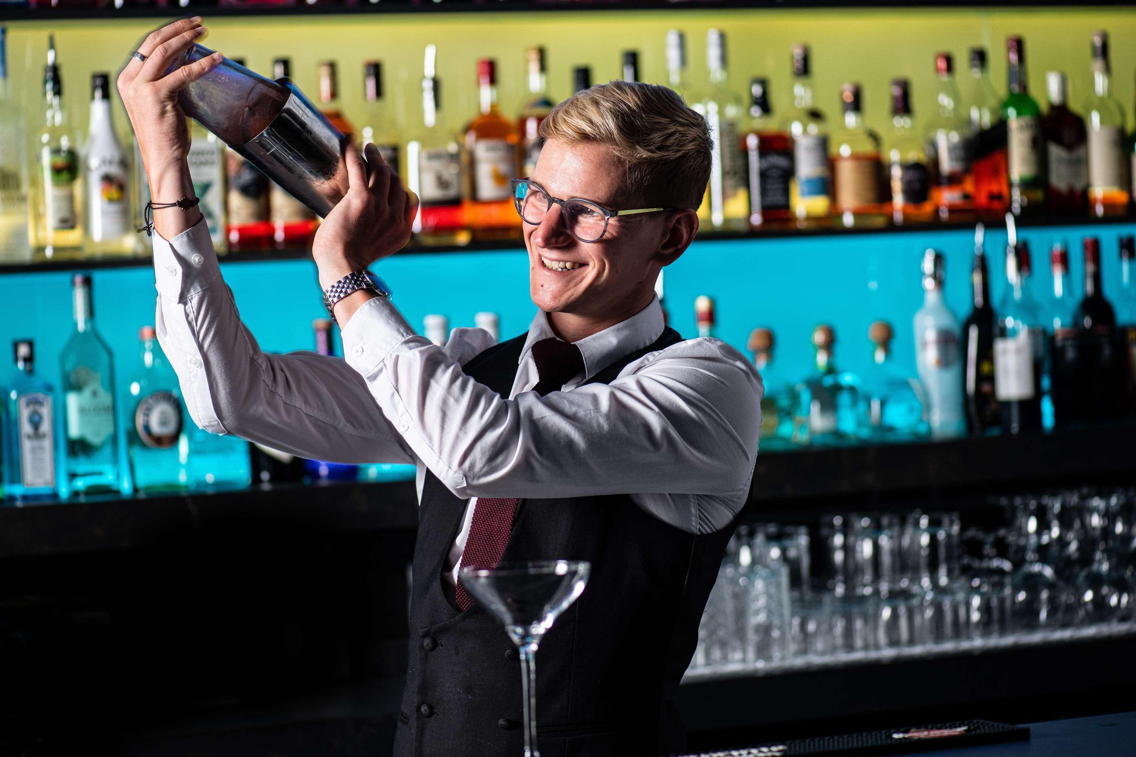 Um bartender servindo uma bebida em um bar, com diversas garrafas ao fundo.