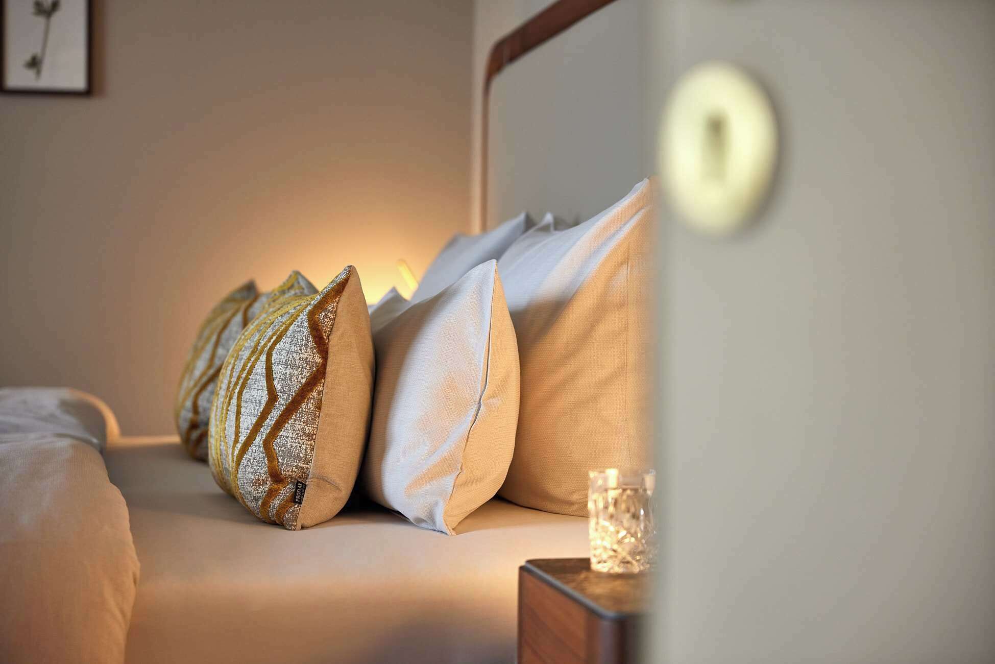 Uma cama bem arrumada com várias almofadas, uma mesinha de cabeceira com um recipiente de vidro e um interruptor de luz na parede.