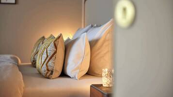 Uma cama bem arrumada com várias almofadas, uma mesinha de cabeceira com um recipiente de vidro e um interruptor de luz na parede.