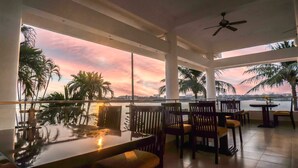 Restaurante com vista para o pôr do sol sobre a água, com mesa e cadeiras, e palmeiras ao fundo.