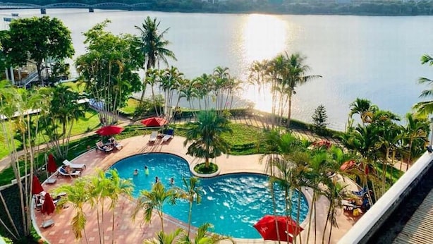 Piscina rodeada por palmeiras e espreguiçadeiras, com vista para um rio e uma ponte ao fundo.
