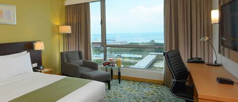 Quarto de hotel com cama, escrivaninha, cadeira, mesinha de cabeceira com abajur, e uma grande janela com vista para o mar.