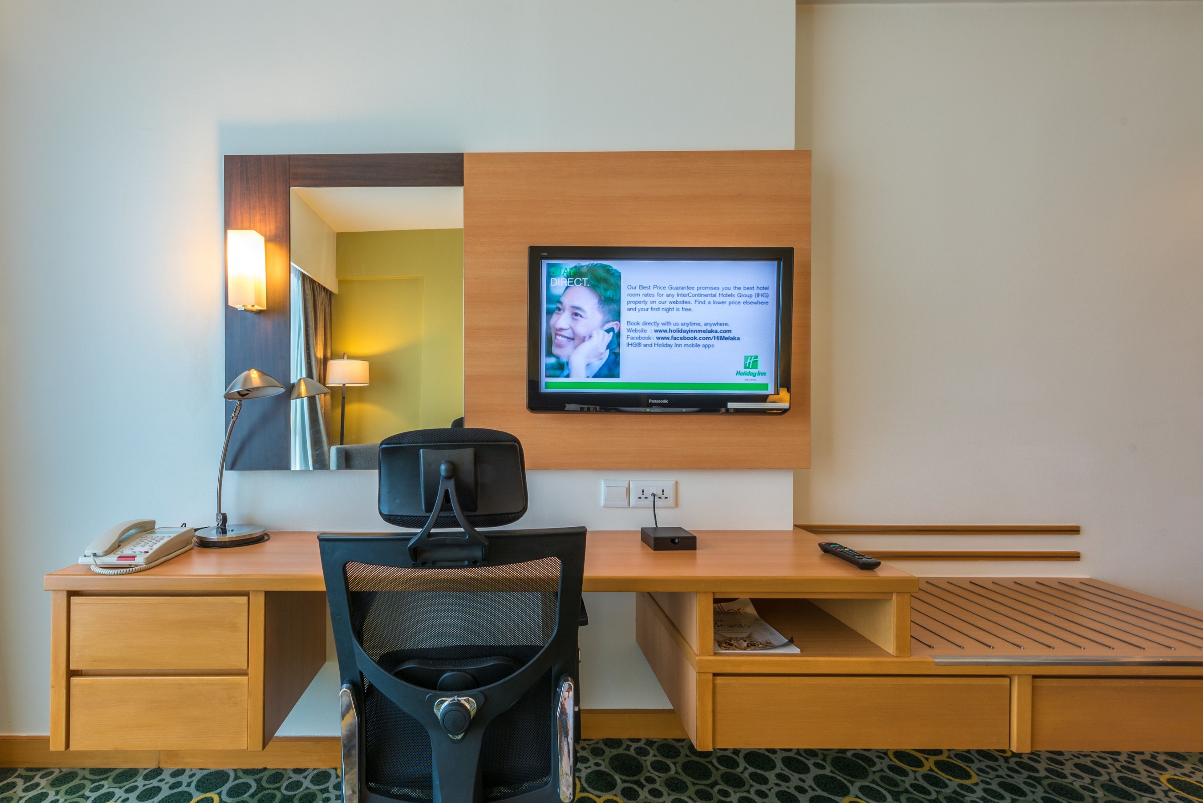 Quarto de hotel com escrivaninha de madeira, uma cadeira, luminária, telefone, televisão fixada na parede e pequena mesa de cabeceira.