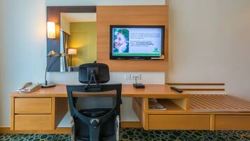 Quarto de hotel com escrivaninha de madeira, uma cadeira, luminária, telefone, televisão fixada na parede e pequena mesa de cabeceira.