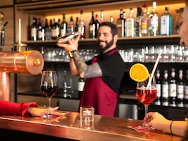 Ambiente de bar com um barman preparando bebidas, uma taça de vinho e um copo de cocktail com uma fatia de laranja.