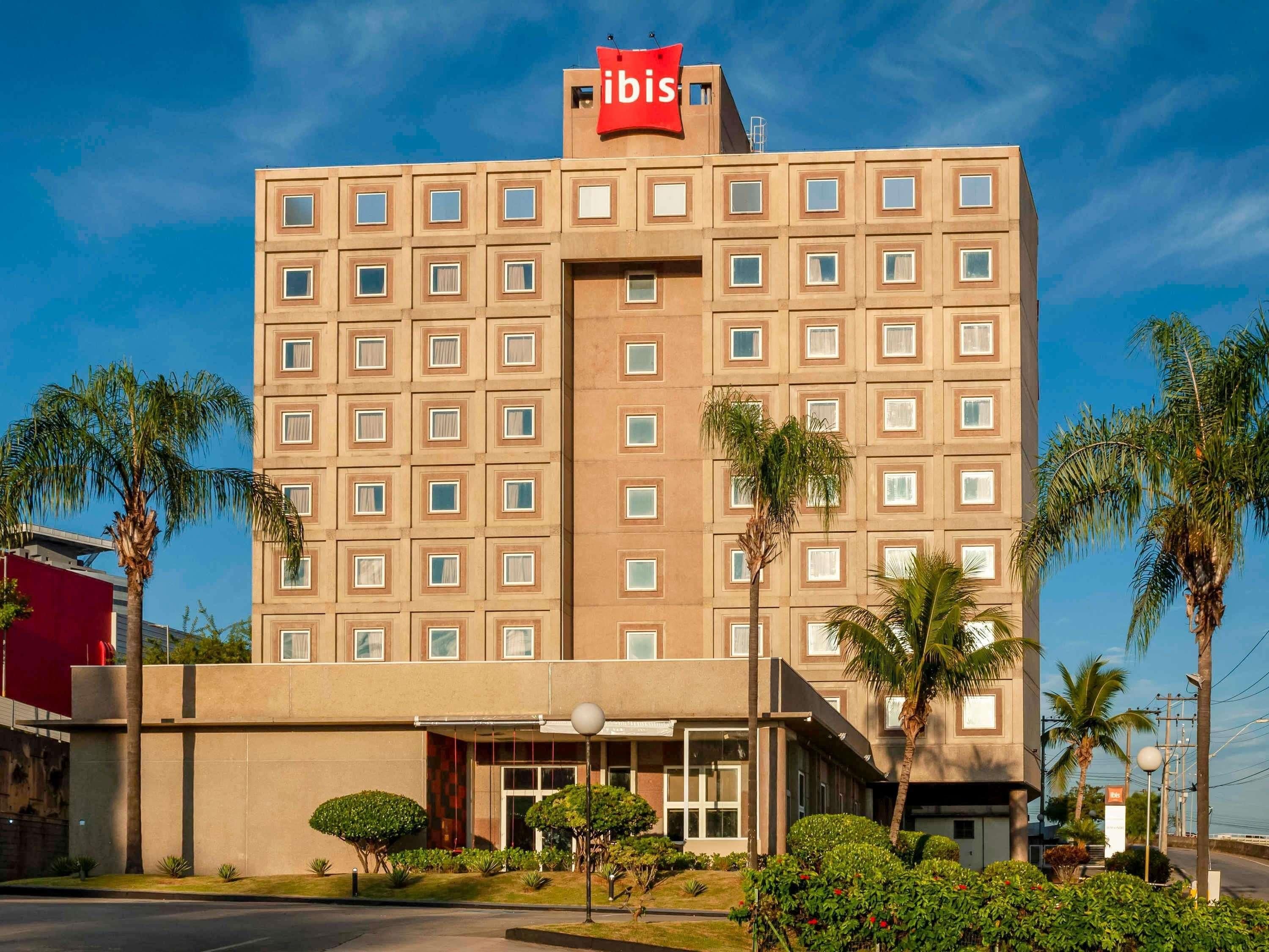 Um hotel com vários andares e um sinal vermelho chamativo da marca 'ibis', apresentando uma fachada simétrica com janelas uniformemente distribuídas e uma entrada central.