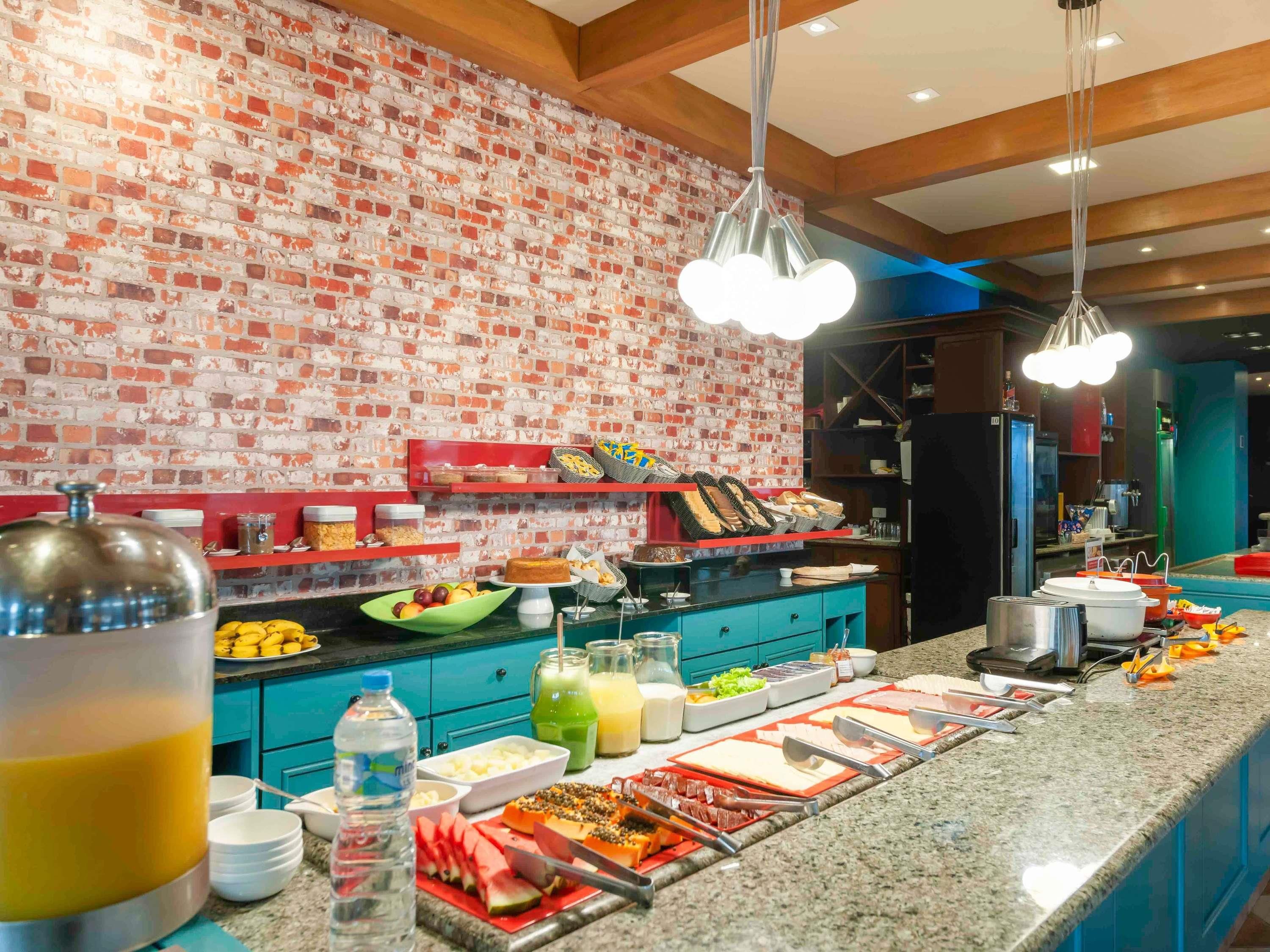 Buffet com variedade de alimentos e bebidas, parede de tijolos e vigas de madeira no teto.