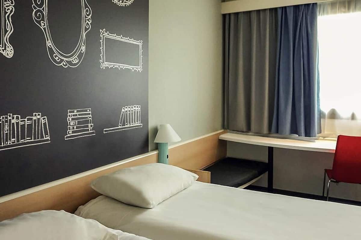 Quarto de hotel com uma cama, uma escrivaninha com cadeira, uma luminária e uma janela com cortinas.