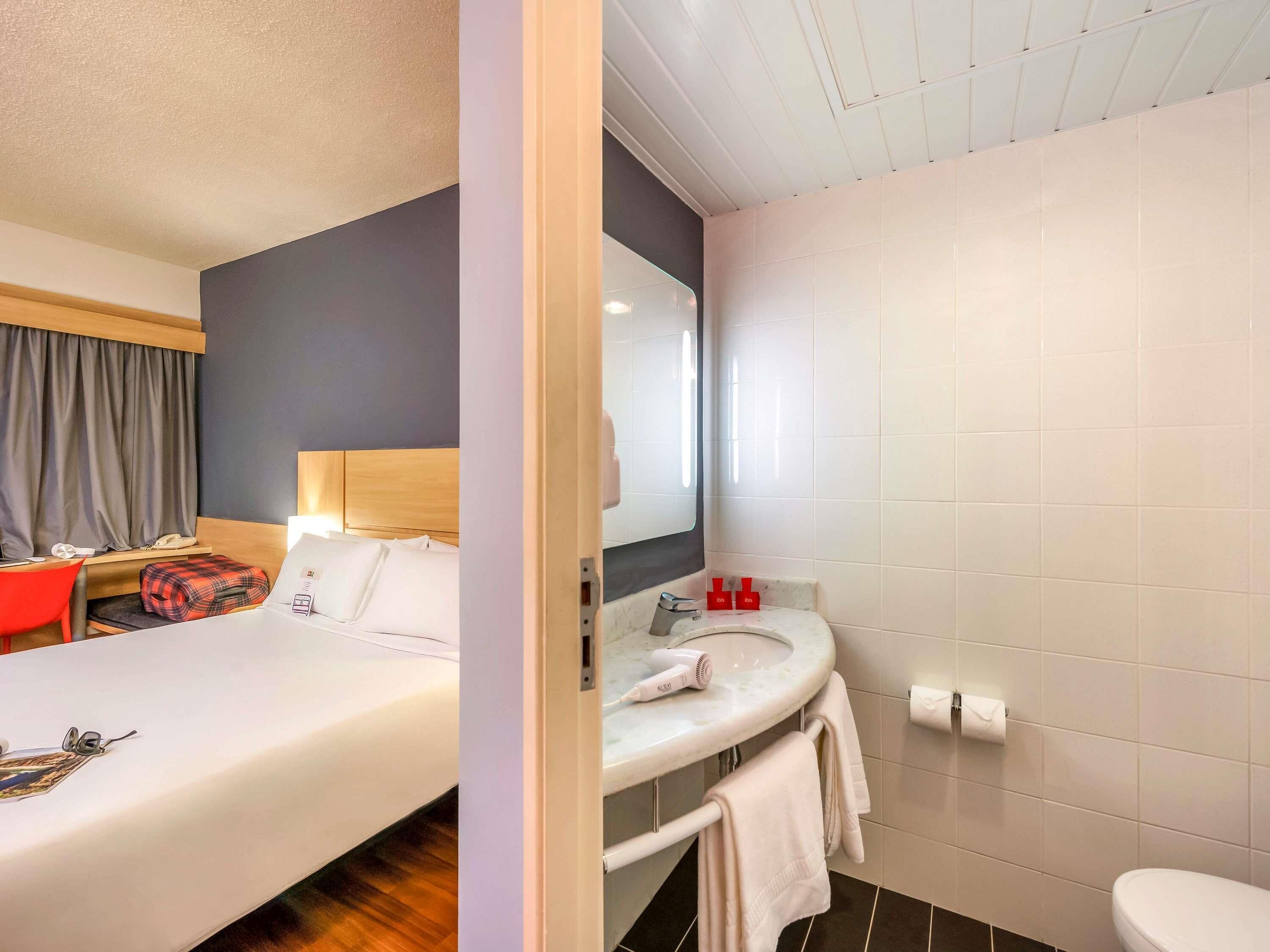 Quarto de hotel com cama, escrivaninha, cadeira, mala, espelho, pia e vaso sanitário.