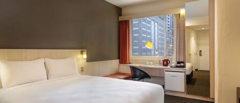 Quarto de hotel com uma cama grande, uma escrivaninha com abajur, uma cafeteira e vista para edifícios altos pela janela.