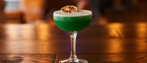 Um cocktail verde num copo com haste, decorado com uma fatia de laranja desidratada, sobre uma mesa de madeira.