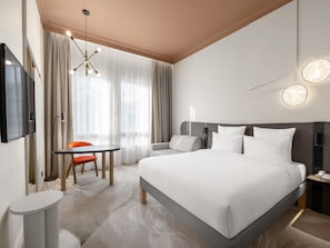 Quarto de hotel com cama, televisão, escrivaninha com cadeira e janela com cortinas.