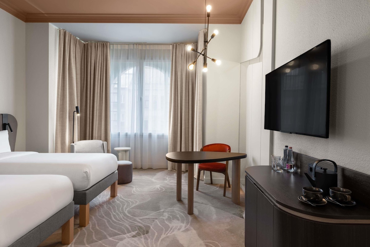 Quarto de hotel com duas camas, uma televisão, uma escrivaninha com duas chávenas, uma cadeira e uma mesa pequena.