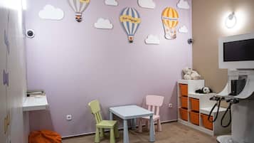 Uma sala de jogos infantil com parede roxa, decorações de balão de ar quente, uma pequena mesa, cadeiras e um console de jogos.