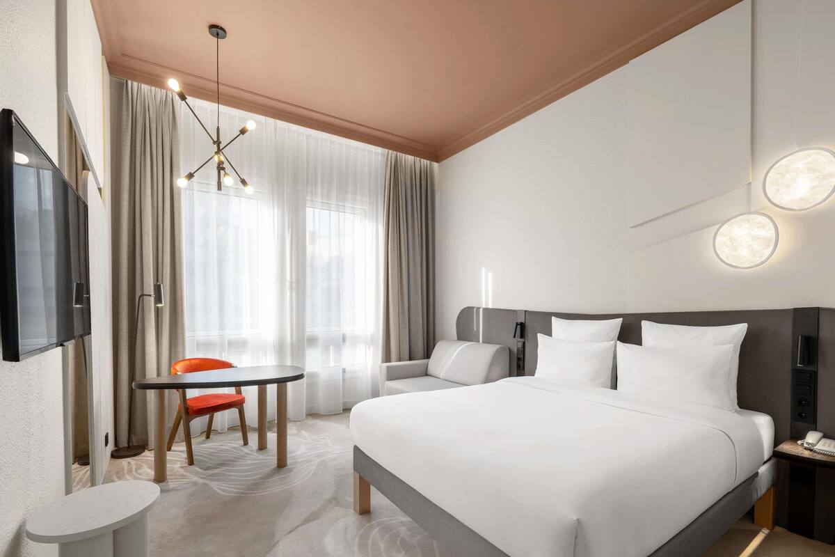 Quarto de hotel com cama, televisão, escrivaninha com cadeira e janela com cortinas.