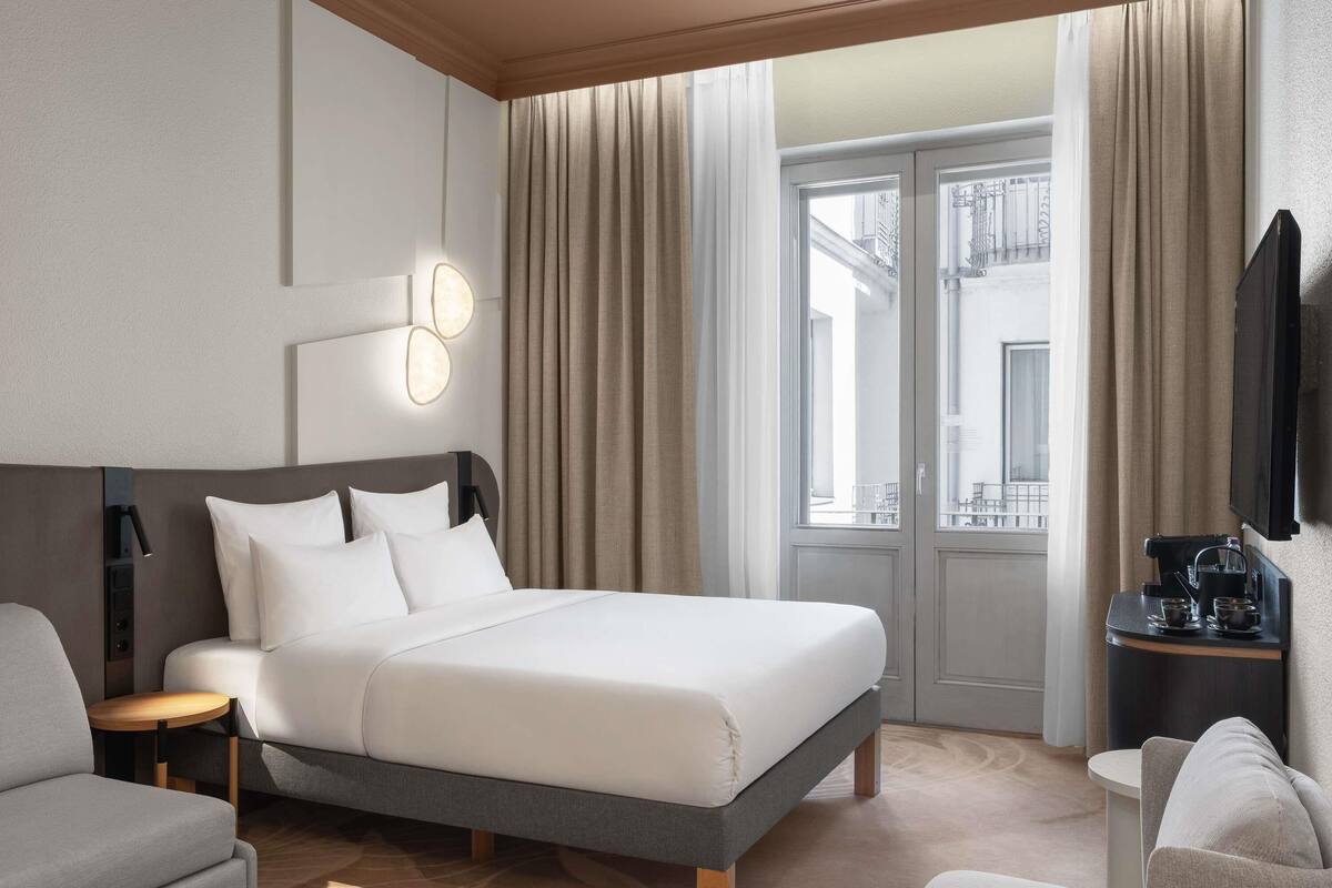Quarto de hotel moderno com uma cama, área de estar com sofá e poltrona, uma pequena mesa com cafeteira e uma TV fixada na parede.