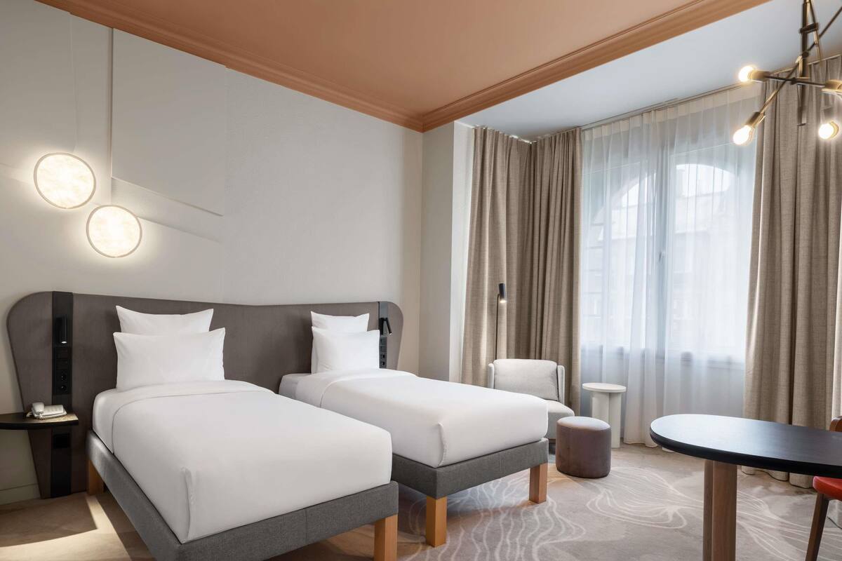 Quarto de hotel com duas camas, uma mesinha, uma cadeira, um abajur e vista da cidade através de cortinas finas.