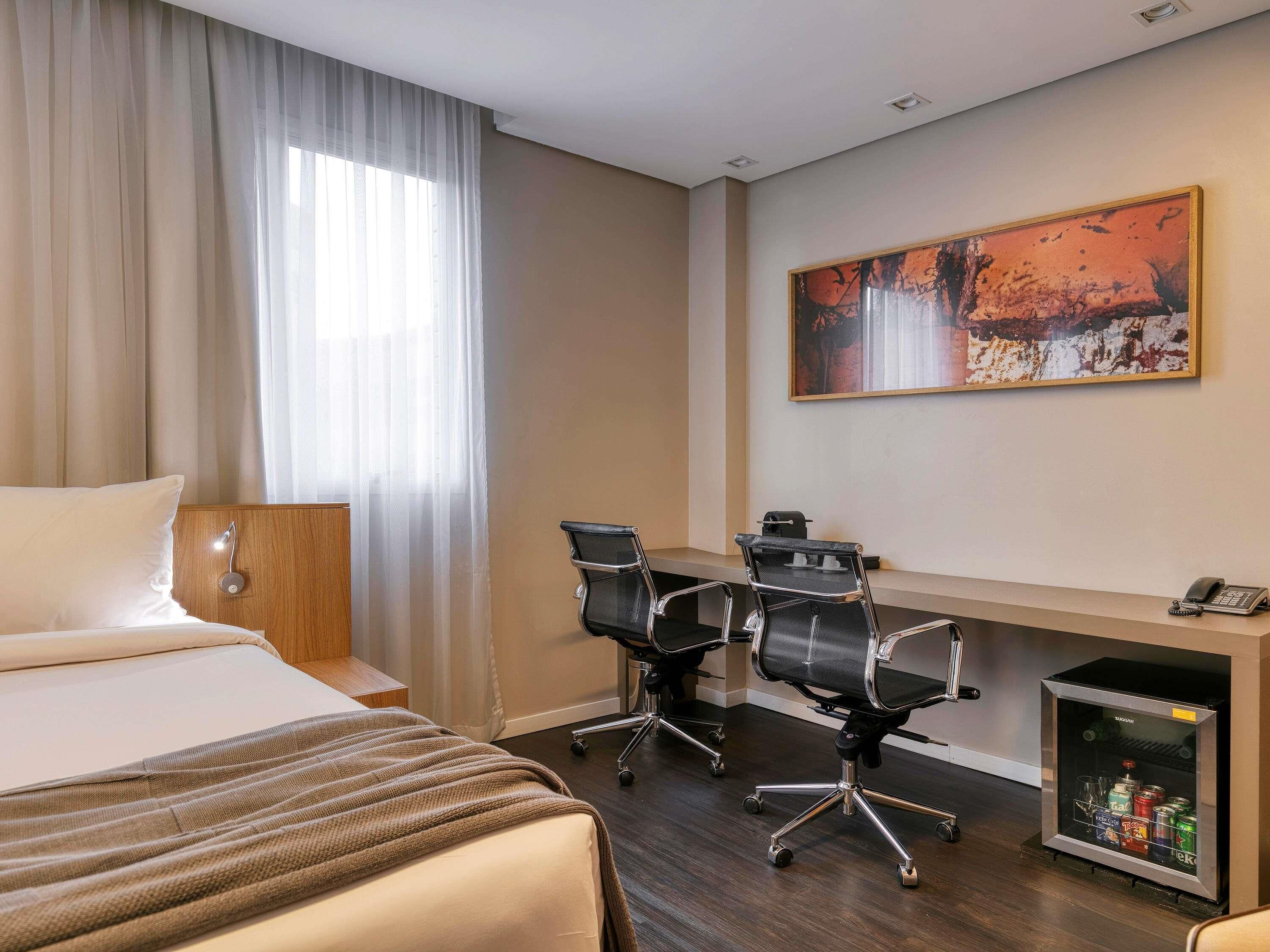Quarto de hotel com uma cama, uma secretária com duas cadeiras, um minibar e um quadro na parede.