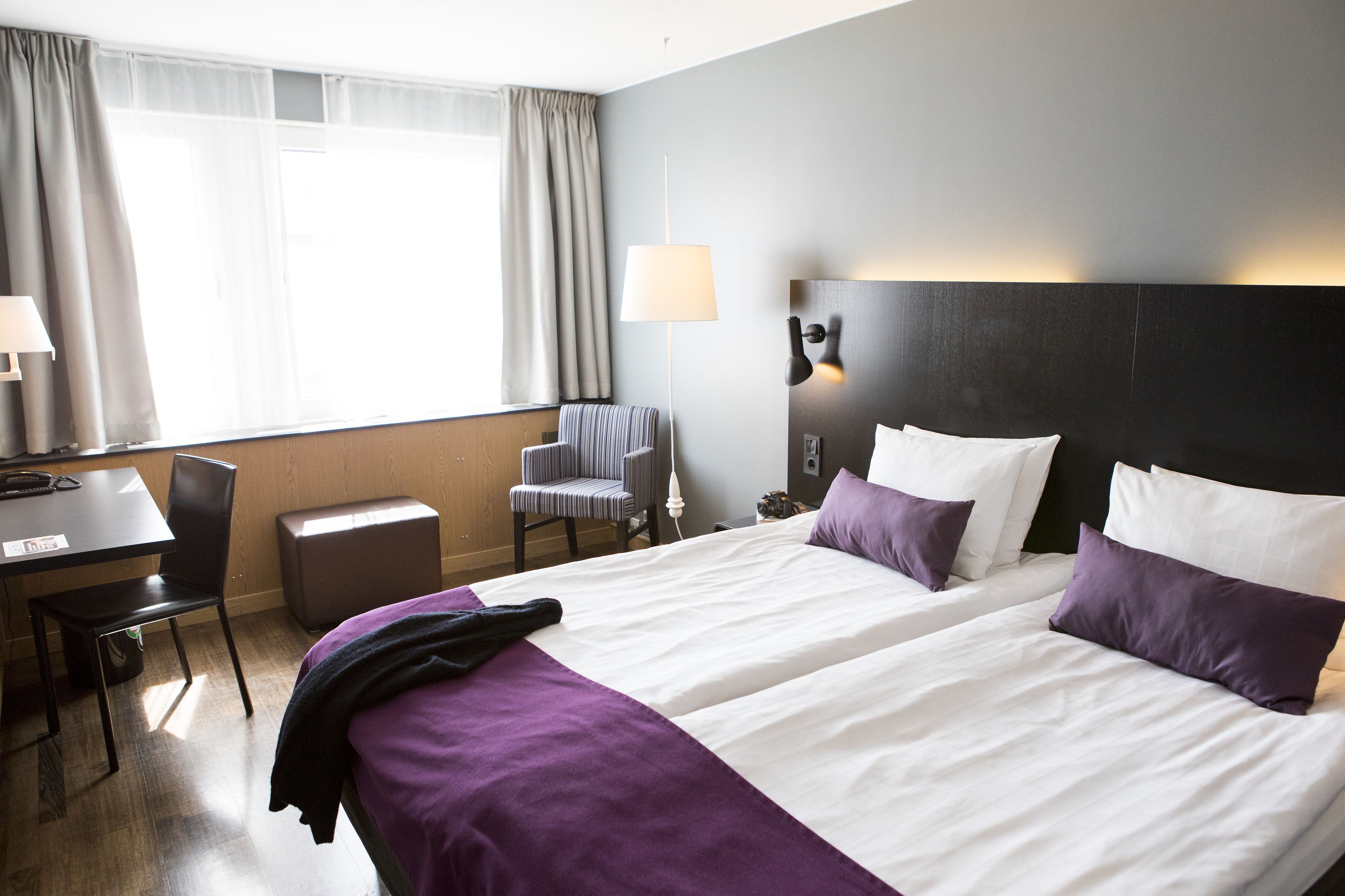 Quarto de hotel com duas camas, uma escrivaninha, uma cadeira, um abajur e uma janela com cortinas.