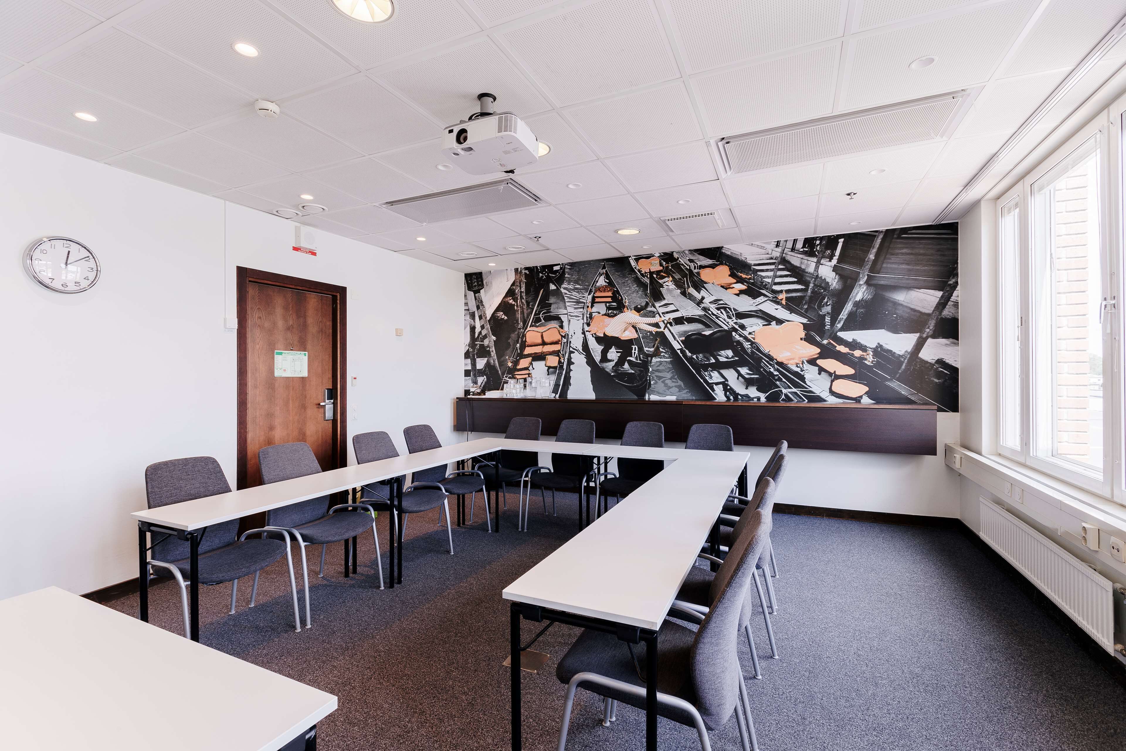 Uma sala de conferências com uma mesa comprida, cadeiras, um relógio, um projetor e um grande mural abstrato na parede.