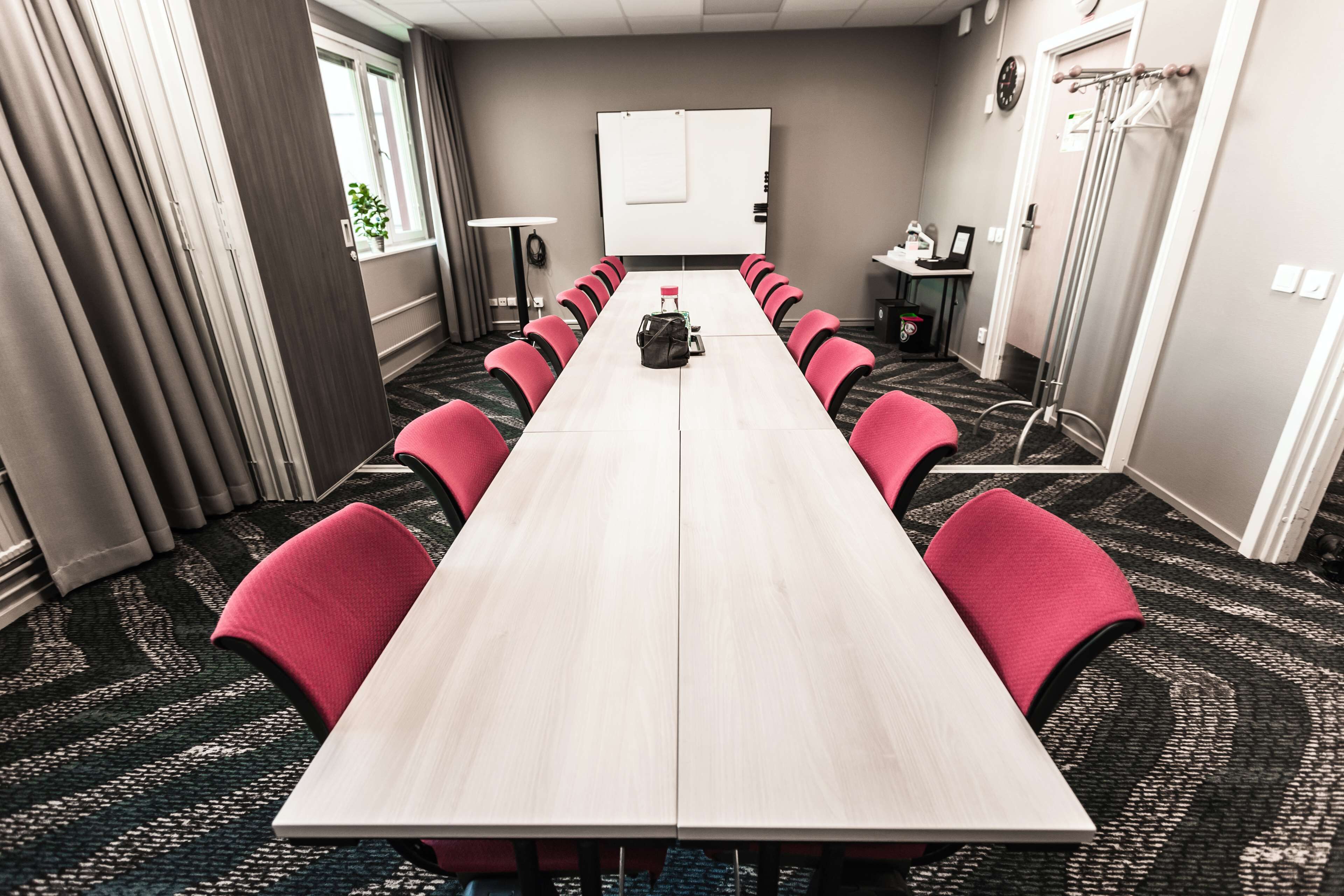 Sala de conferências com uma mesa comprida, cadeiras vermelhas, um quadro branco e um relógio na parede.