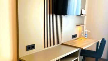 Quarto moderno com TV integrada, escrivaninha de madeira, cadeira e prateleira pequena.