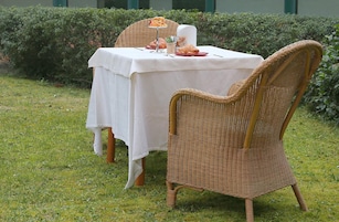 Mesa posta ao ar livre com cadeiras de vime, toalha de mesa branca e uma pequena mesa com comida e bebidas.