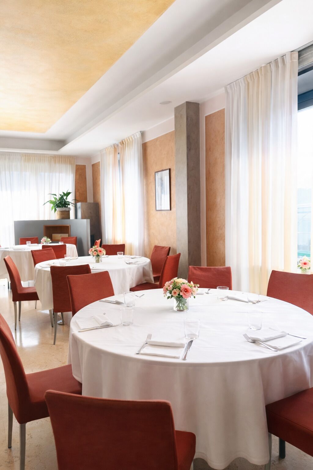 Uma área de refeições com mesas redondas, cadeiras vermelhas e toalhas de mesa brancas.