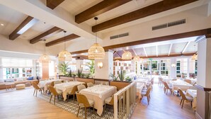 Um restaurante amplo com pé direito alto, vigas de madeira e luminárias pendentes. Mesas montadas para refeições, com plantas e áreas de estar visíveis.