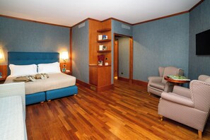 Quarto de hotel com uma cama grande, área de estar com duas poltronas, aparador de madeira e televisão de tela plana na parede.