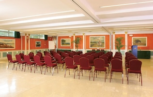 Uma sala de conferências com fileiras de cadeiras vermelhas, um púlpito e quadros nas paredes.