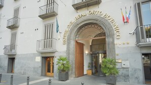 A entrada do Hotel del Real Ortiz Botânico, com um arco de pedra, plantas em vasos e bandeiras.