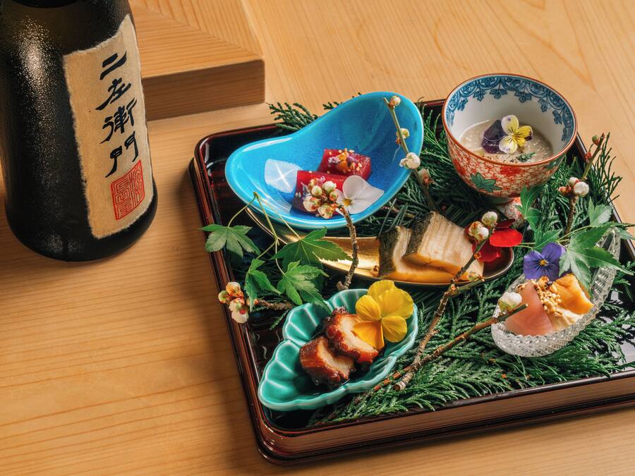 Uma bandeja com comida e bebidas, incluindo uma garrafa com caracteres japoneses, sobre uma mesa de madeira.