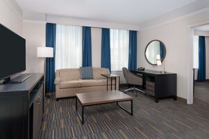 Quarto de hotel com sofá, escrivaninha, cadeira, televisão e um espelho grande.