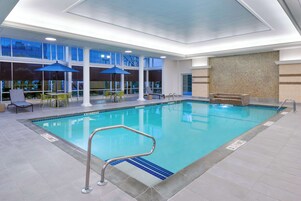 Piscina interior com água azul cristalina, escada de metal e área de estar com mesas e cadeiras.