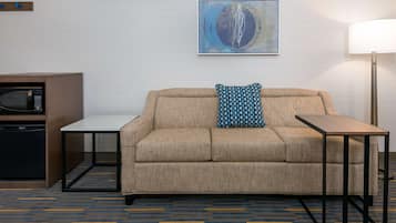 Quarto de hotel com um sofá bege, uma almofada estampada, uma mesinha, um micro-ondas e uma luminária.